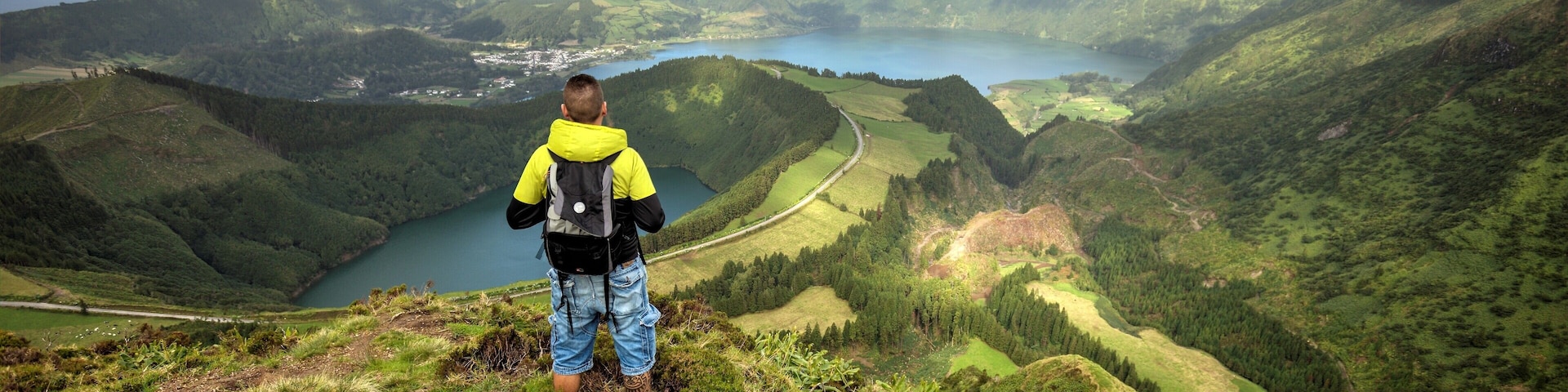 Uma pessoa em pé numa colina gramada, com vista para um vale com um lago e estradas sinuosas.
