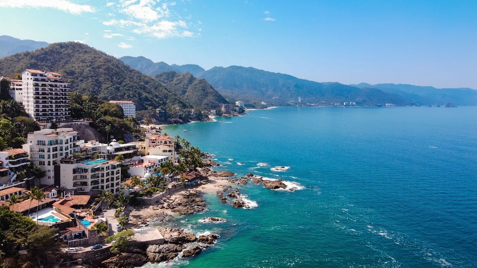 Uma cidade costeira com edifícios, praia, linha costeira rochosa e montanhas ao fundo.