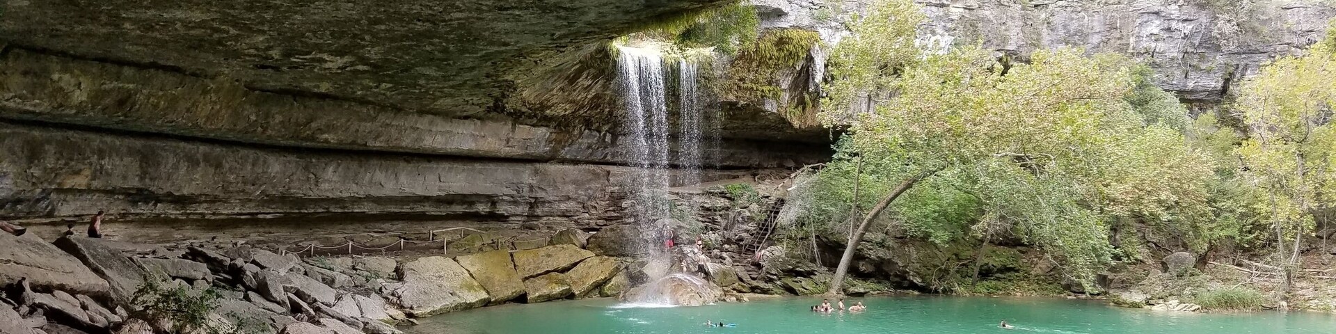 Uma cascata sob uma saliência rochosa, com águas cristalinas e pessoas a nadar.