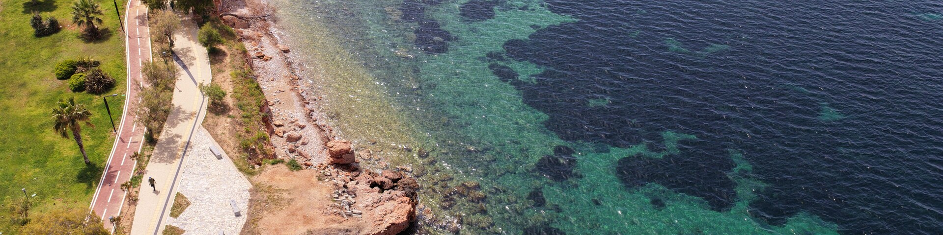 Paisagem costeira com mar azul límpido, linha de costa rochosa e calçada pavimentada com bancos.