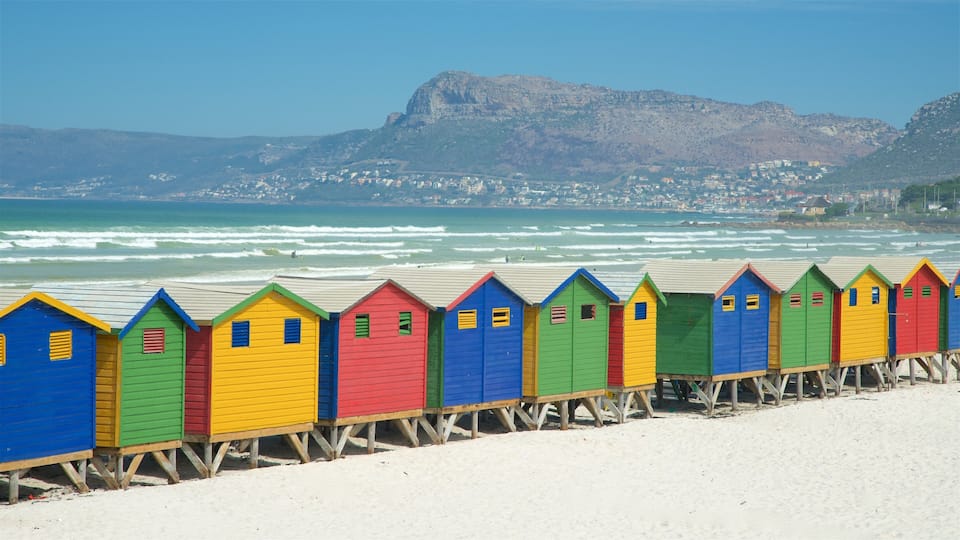 Uma fileira de cabanas coloridas sobre palafitas, ao longo de uma praia de areia, com ondas ao fundo e uma cordilheira.