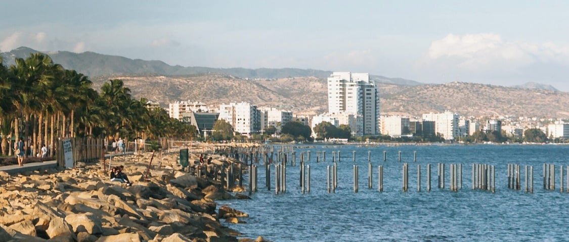 Uma costa rochosa com um cais que se estende sobre águas calmas, palmeiras e uma paisagem urbana ao fundo.