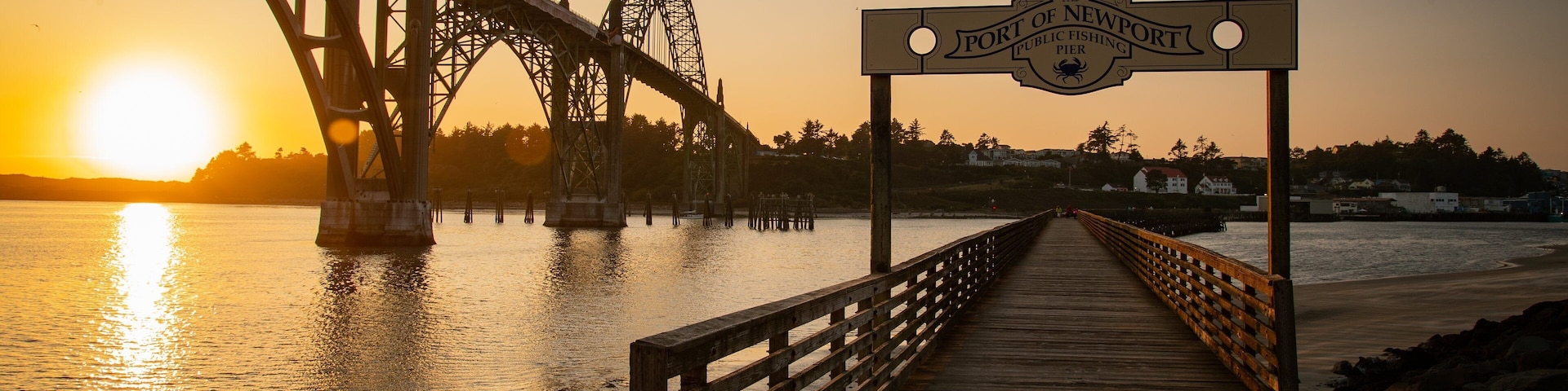 Um pôr do sol sobre uma ponte, um cais de madeira e uma placa com a inscrição "The Port of Newport Public Fishing Pier".