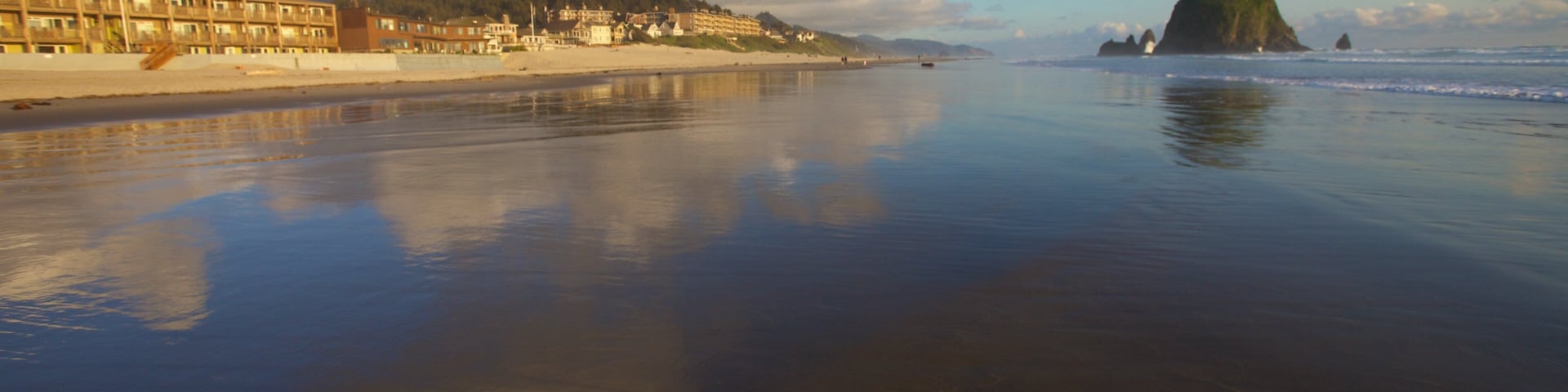 Uma praia com mar calmo, uma grande formação rochosa e edifícios ao longo da costa.