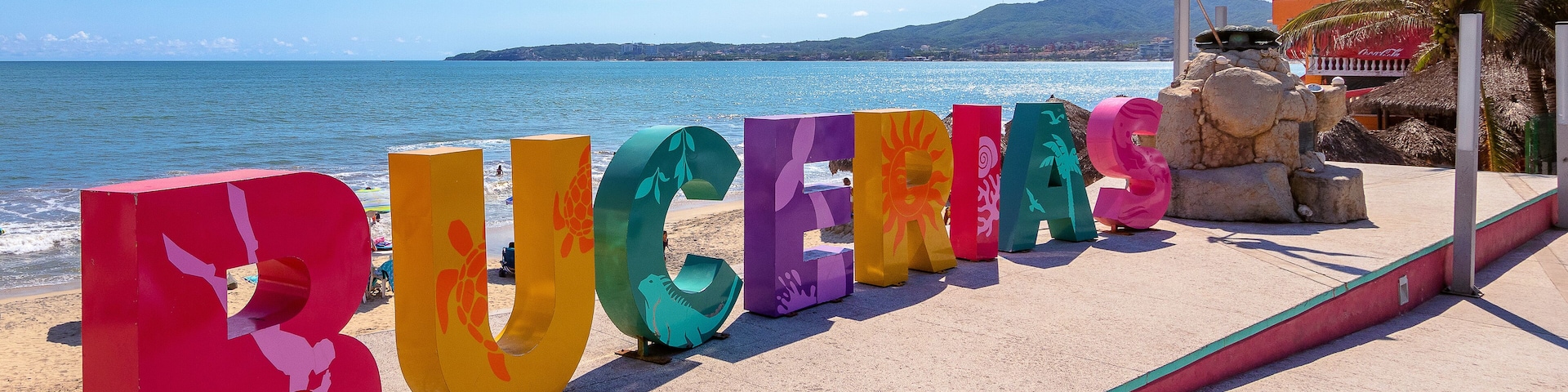 Large colorful letters spelling "BUCHERAS" on a beach with the ocean and mountains in the background.