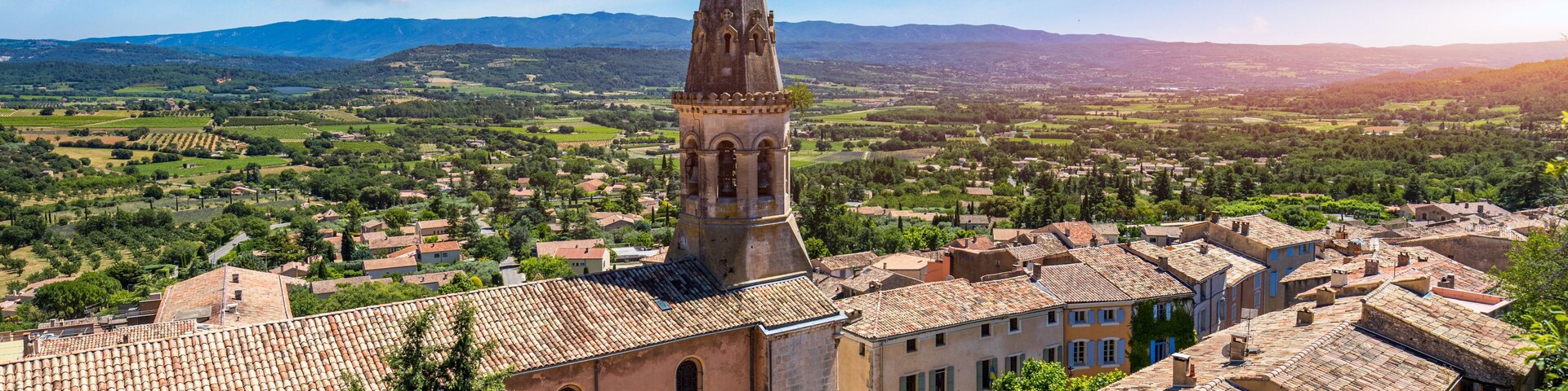 Uma cidade com uma torre de igreja imponente, edifícios com telhados de terracota e uma paisagem montanhosa ao fundo.