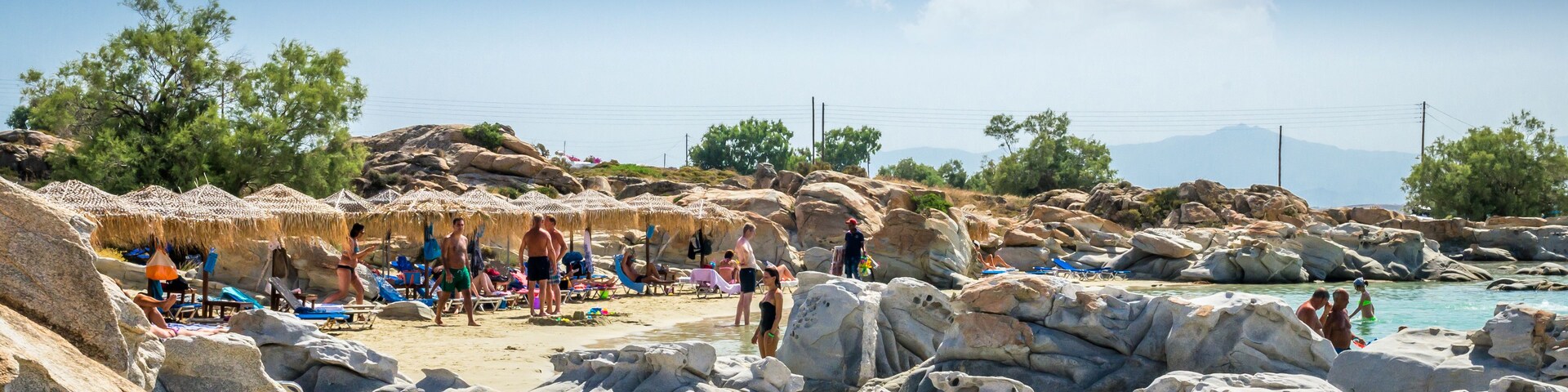 Uma praia rochosa com guarda-sóis de palha e pessoas tomando sol.