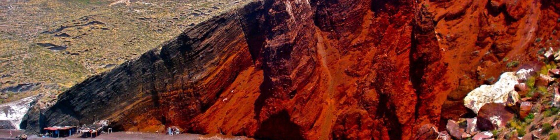 Paisagem costeira com falésias avermelhadas, águas azul-claras e uma praia com pessoas.
