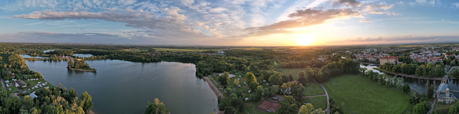 Vista panorâmica de uma cidade à beira do lago ao pôr do sol, com um lago extenso, vegetação ao redor e uma combinação de edifícios residenciais e comerciais.