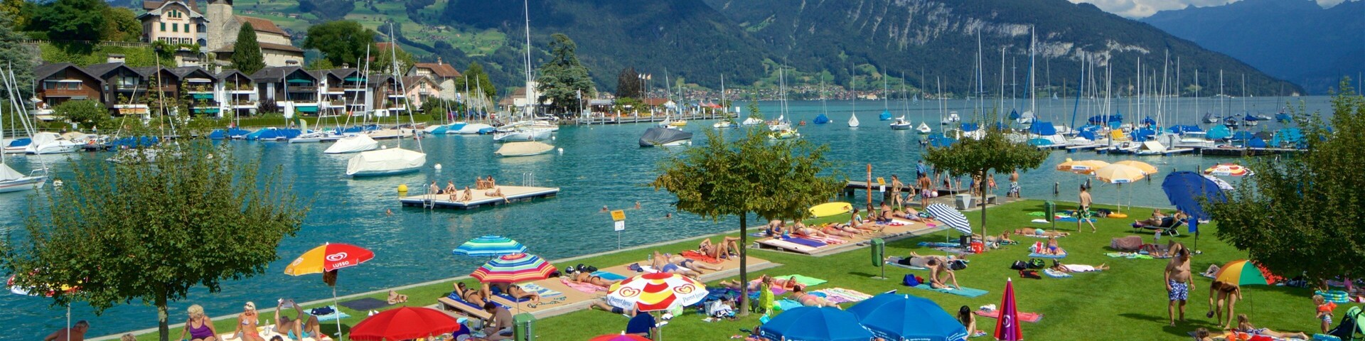 Uma marina à beira do lago com barcos, pessoas tomando sol e montanhas ao fundo.
