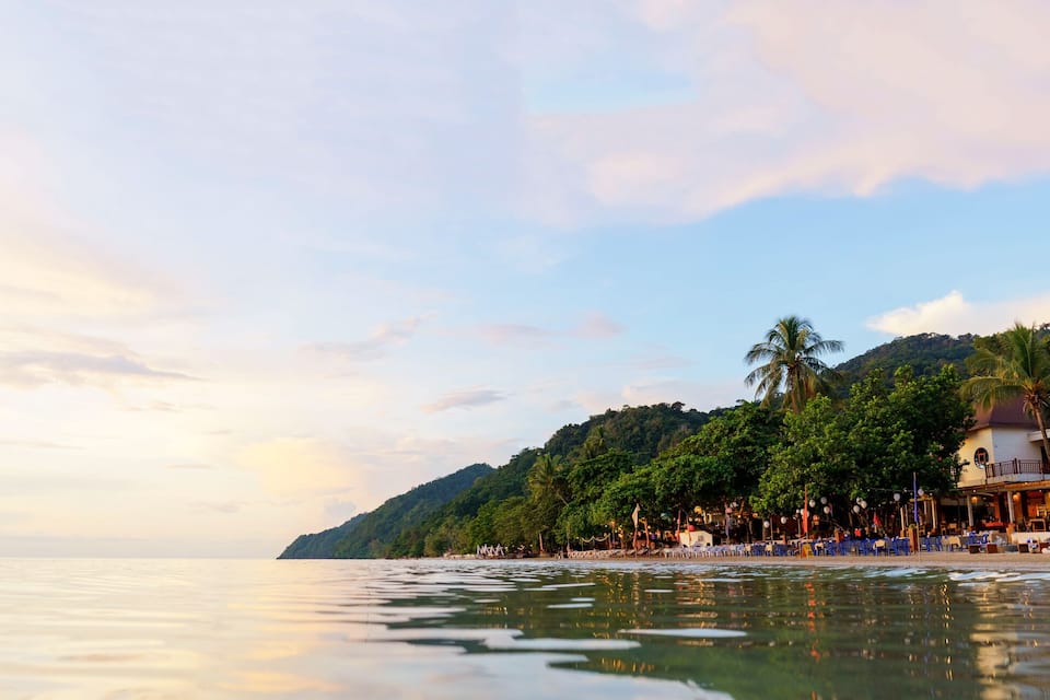 Uma praia com águas calmas, um edifício com telhado de palha e vegetação exuberante numa encosta.