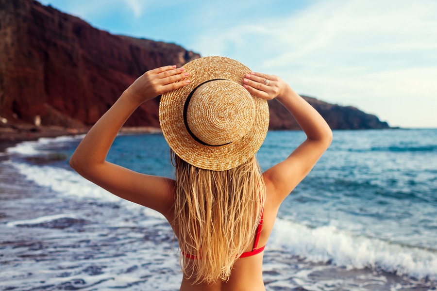 Uma pessoa usando chapéu de palha e biquíni vermelho, em pé numa praia com ondas e um penhasco rochoso ao fundo.