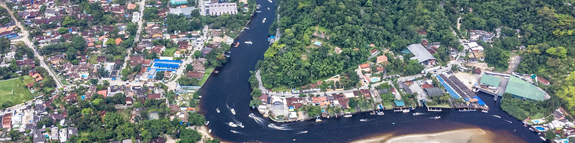 Vista aérea de uma cidade costeira com um rio, floresta densa e uma praia com guarda-sóis.