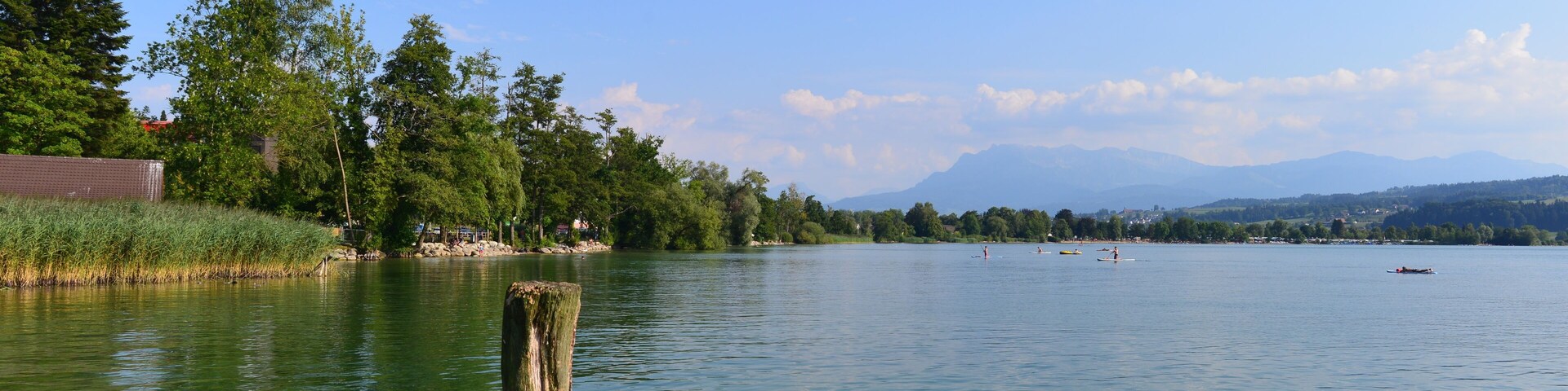Um lago tranquilo com um poste de madeira em primeiro plano, rodeado por árvores e montanhas ao fundo.