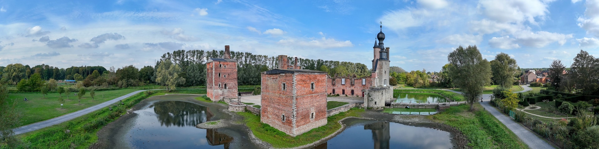 Um edifício histórico de tijolos com uma torre, rodeado por um fosso e vegetação.