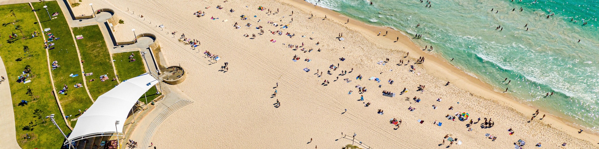 Vista aérea de uma praia com pessoas, guarda-sóis e uma grande estrutura de tenda branca.