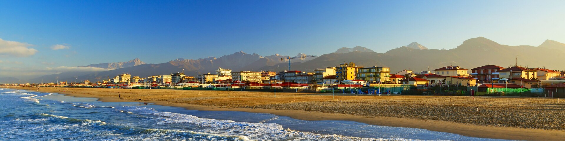 Uma praia com ondas, edifícios e montanhas ao fundo.