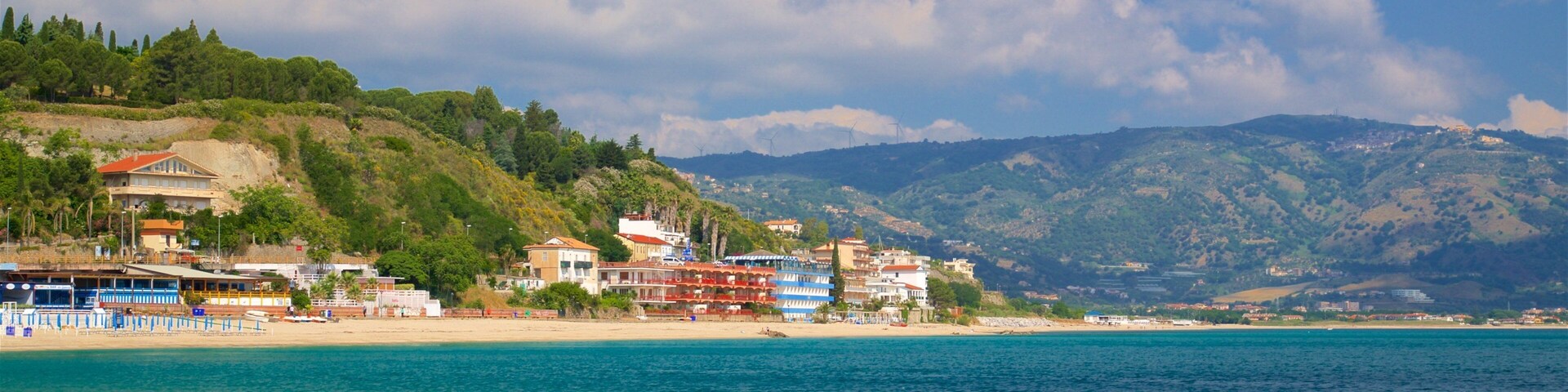 Uma vila costeira com edifícios ao longo da praia, mar azul cristalino e montanhas ao fundo.