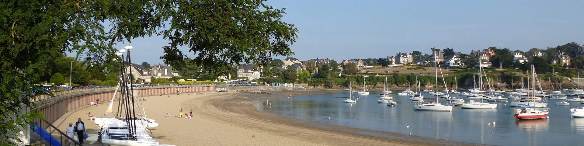 Uma praia com barcos, areal e casas ao fundo.
