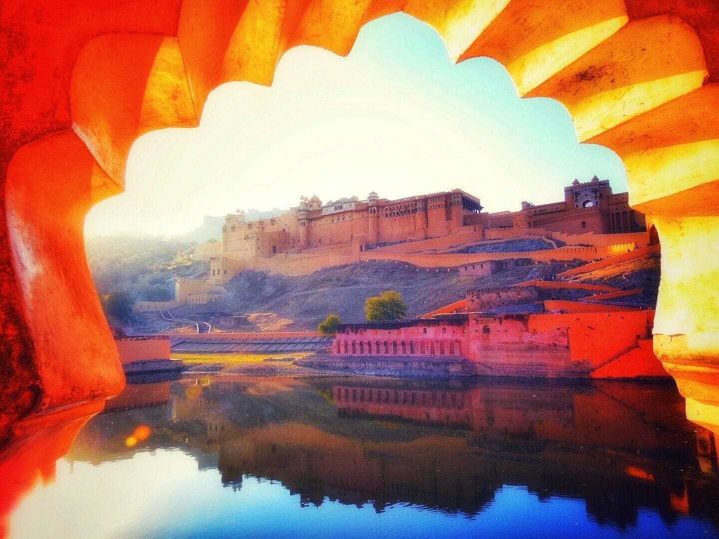 Uma fortaleza histórica refletida na água, cercada por uma paisagem avermelhada e alaranjada.