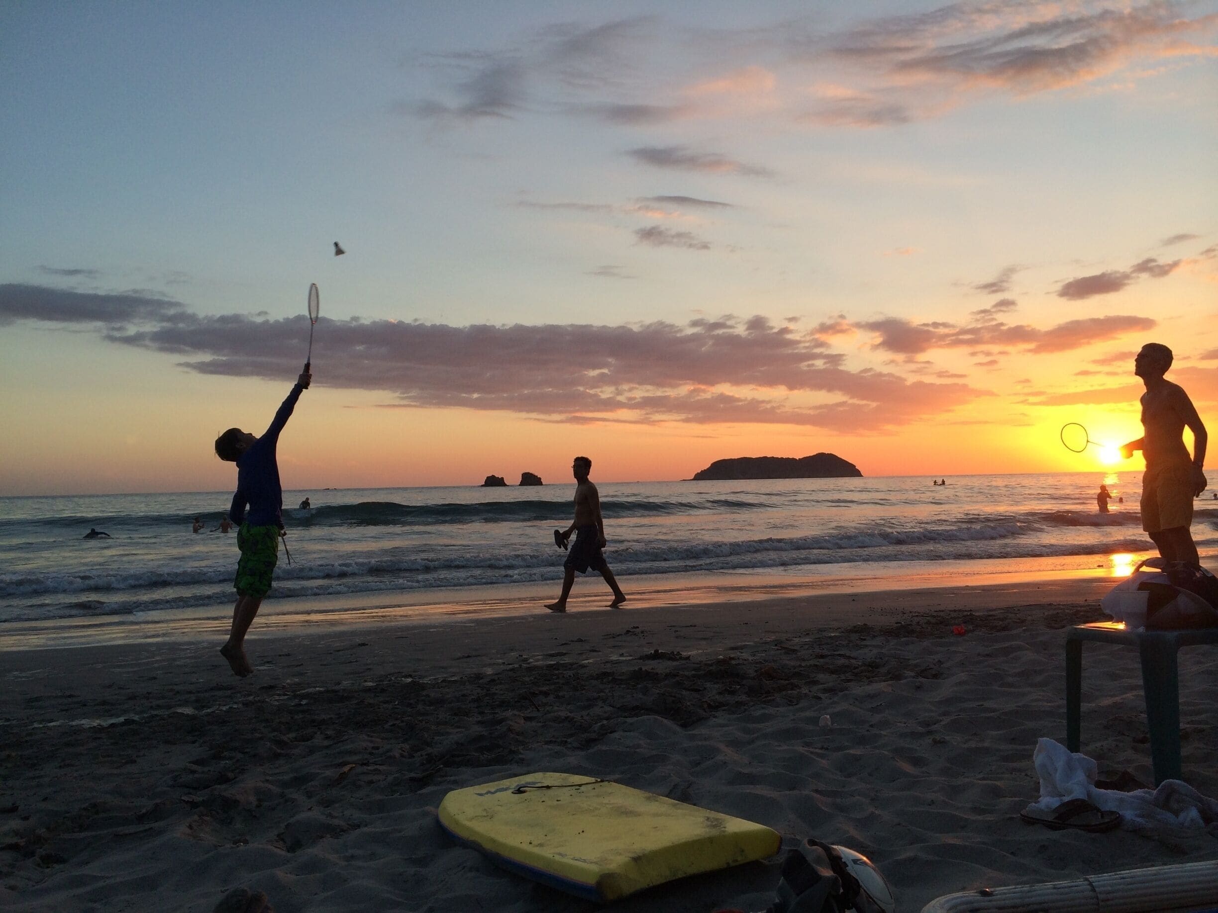 Praia ao pôr do sol com pessoas brincando, ondas e uma ilha distante.