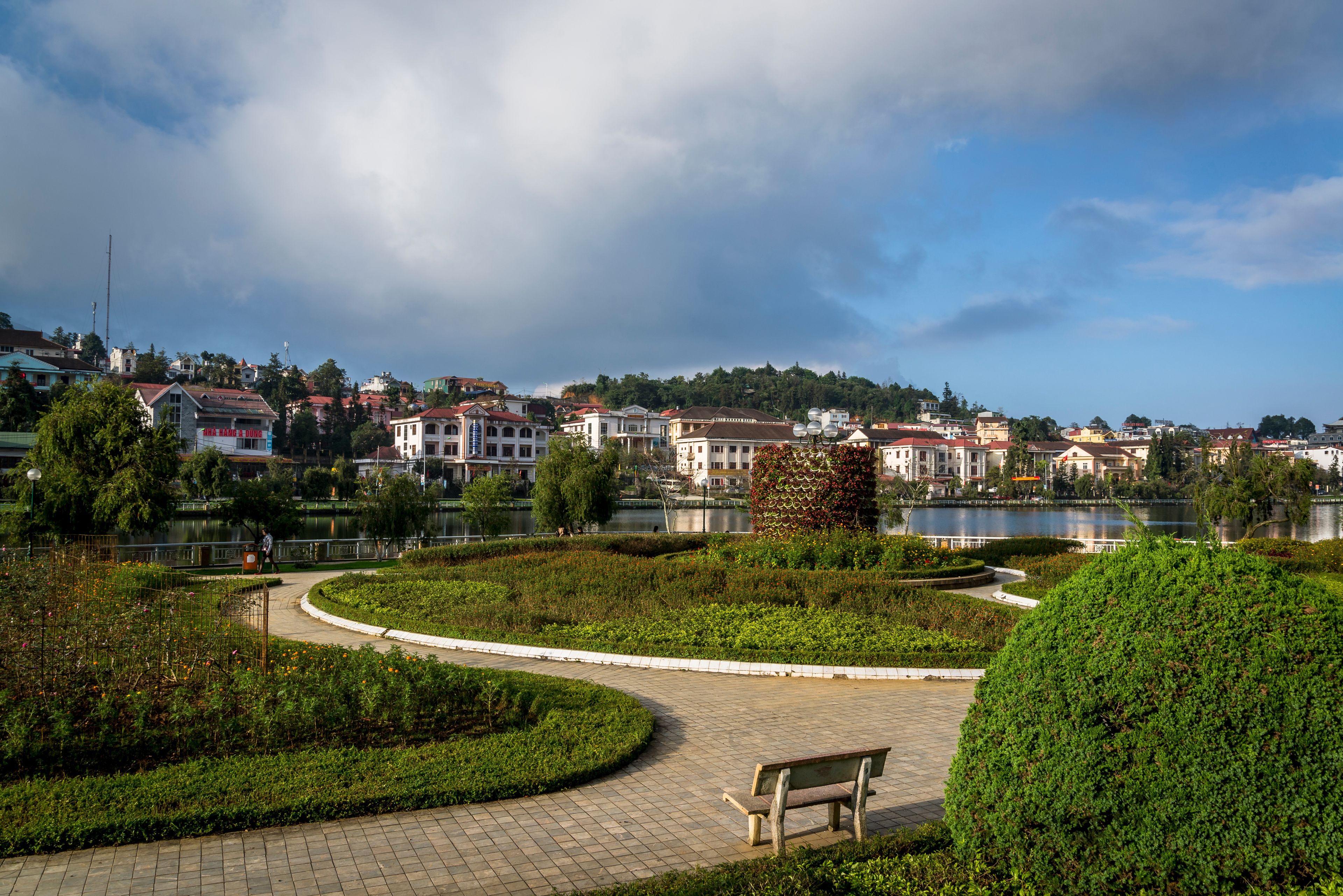 Um parque com um caminho sinuoso, um banco e um lago com edifícios ao fundo.