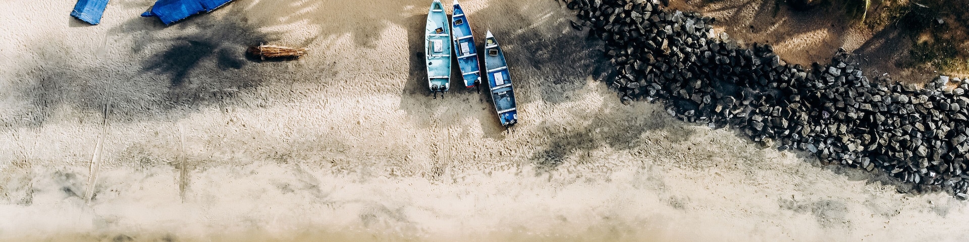 Uma praia arenosa com barcos, palmeiras e um trecho de costa rochosa.