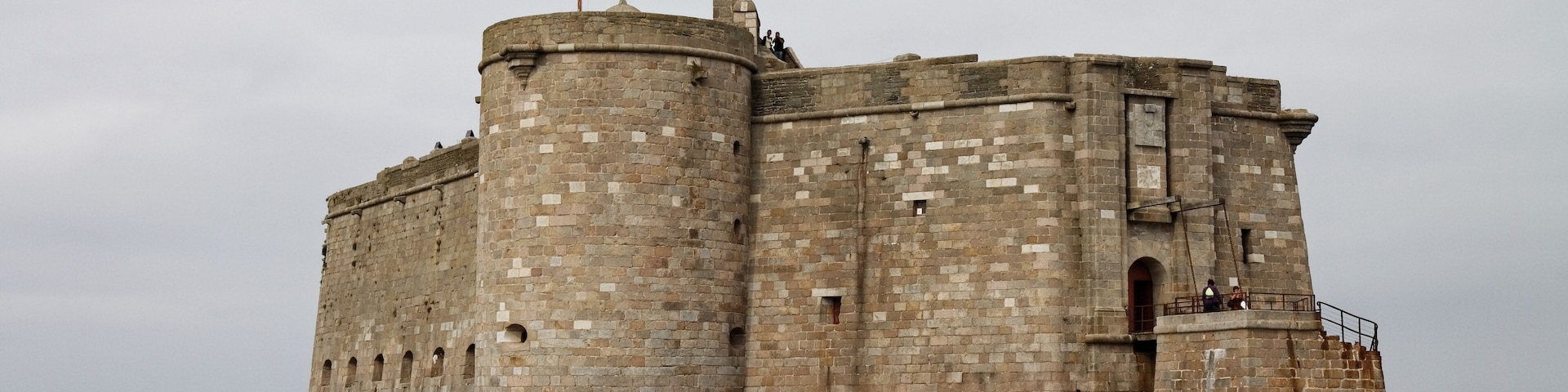 Um castelo de pedra imponente à beira-mar, com uma bandeira no topo.