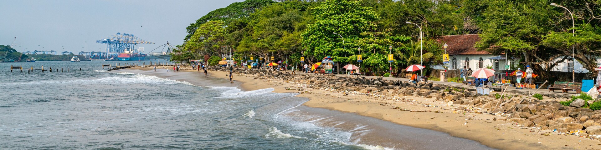 Uma praia costeira com ondas, um litoral rochoso e um porto distante com guindastes.