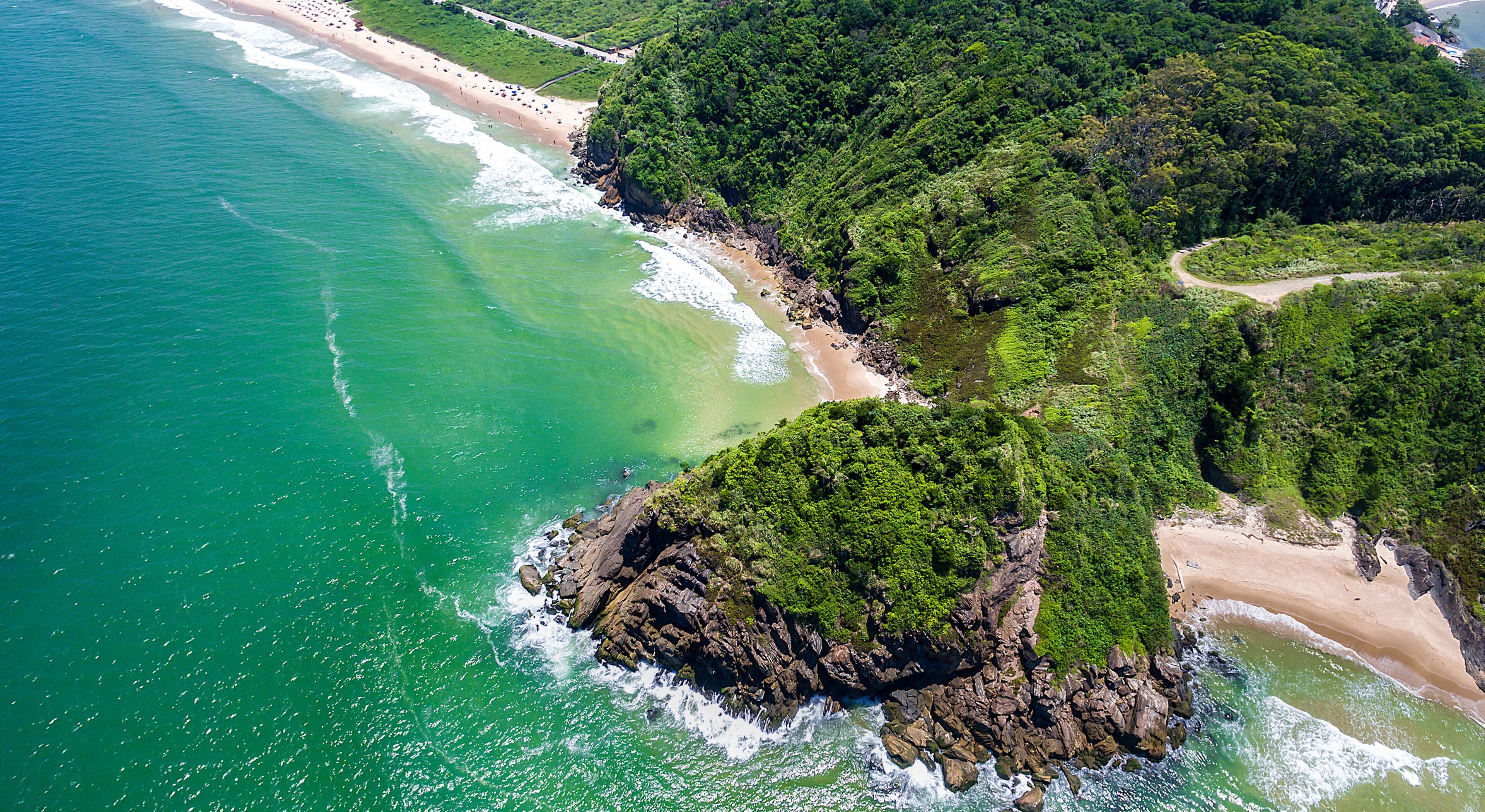 Paisagem costeira com praia, afloramentos rochosos e vegetação densa.