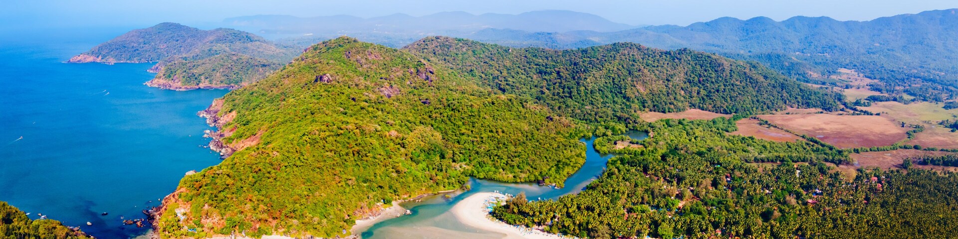 Paisagem costeira com um rio desembocando no mar, cercada por colinas verdes e uma praia com guarda-sóis.