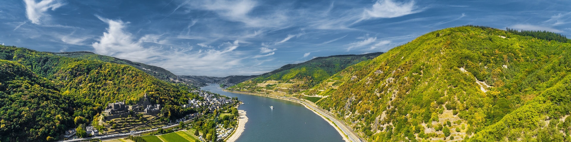 Um rio serpenteando por um vale, com colinas verdejantes, uma pequena cidade com casas e campos, e um céu azul límpido.