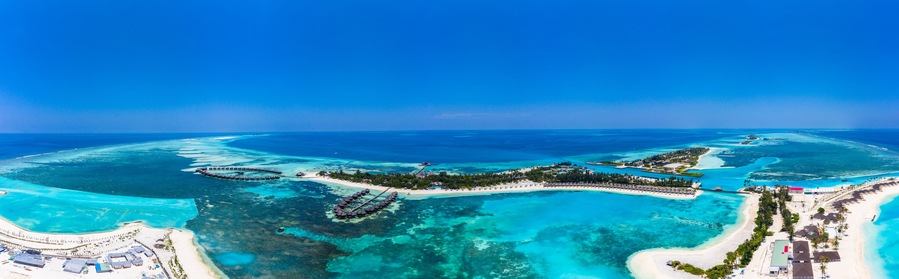 Vista aérea de uma ilha tropical com águas turquesa cristalinas, bangalôs sobre a água e uma praia de areia branca.
