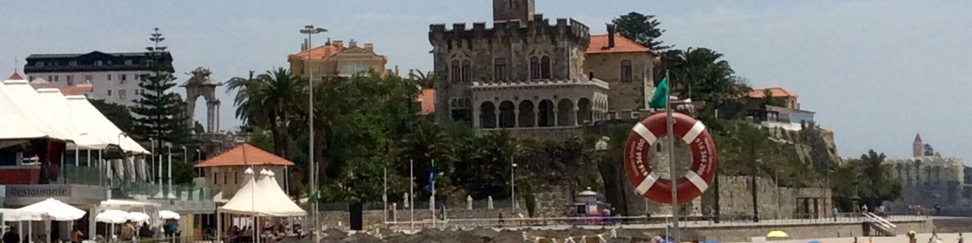 Uma praia com pessoas tomando sol, guarda-sóis e um edifício semelhante a um castelo ao fundo.