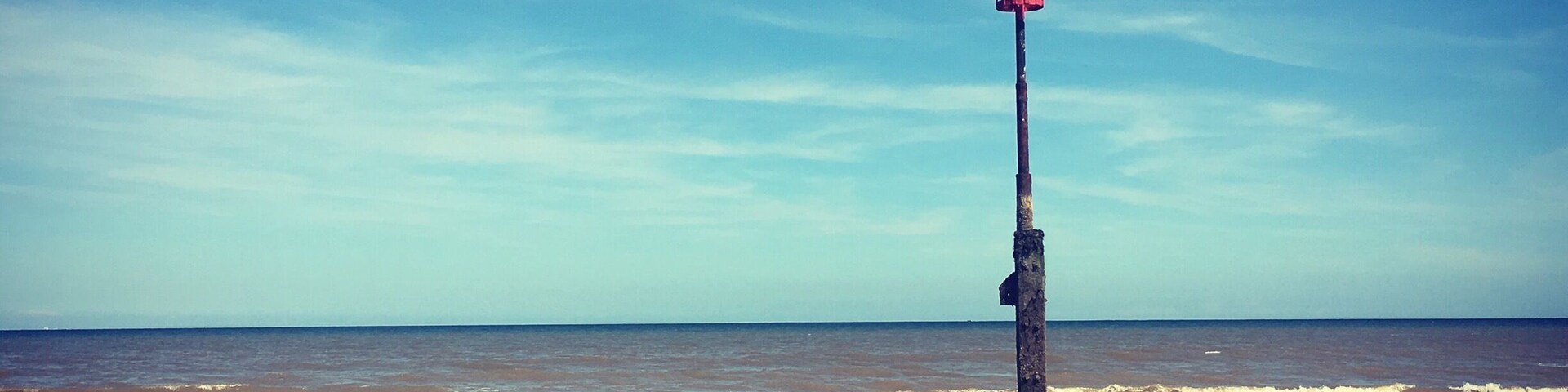Uma praia com um poste de madeira e uma placa vermelha, ondas ao fundo e um céu azul claro.