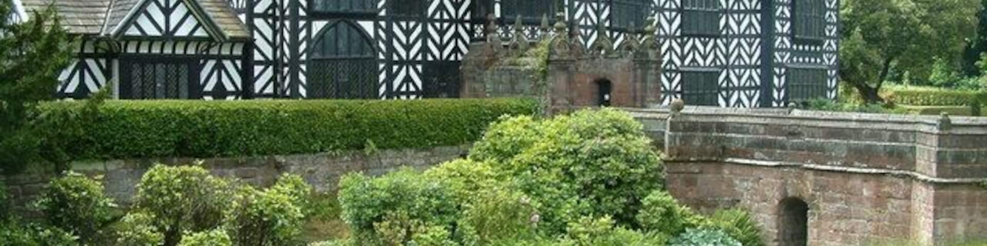 Um edifício em estilo Tudor com estrutura de madeira branca e base de tijolos, rodeado por vegetação exuberante e um muro de pedra.