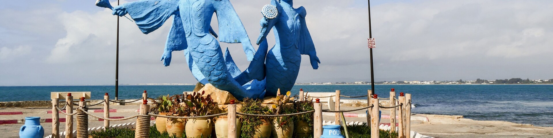 Estátua azul de uma sereia com um homem, rodeados por plantas em vasos e uma cerca, junto ao mar.
