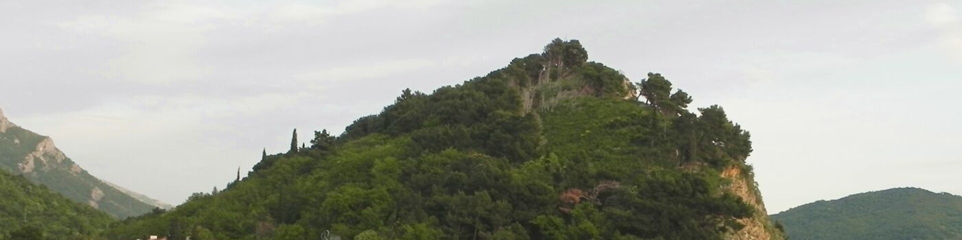 Uma área costeira com uma colina coberta por vegetação, uma praia com pessoas e edifícios ao fundo.