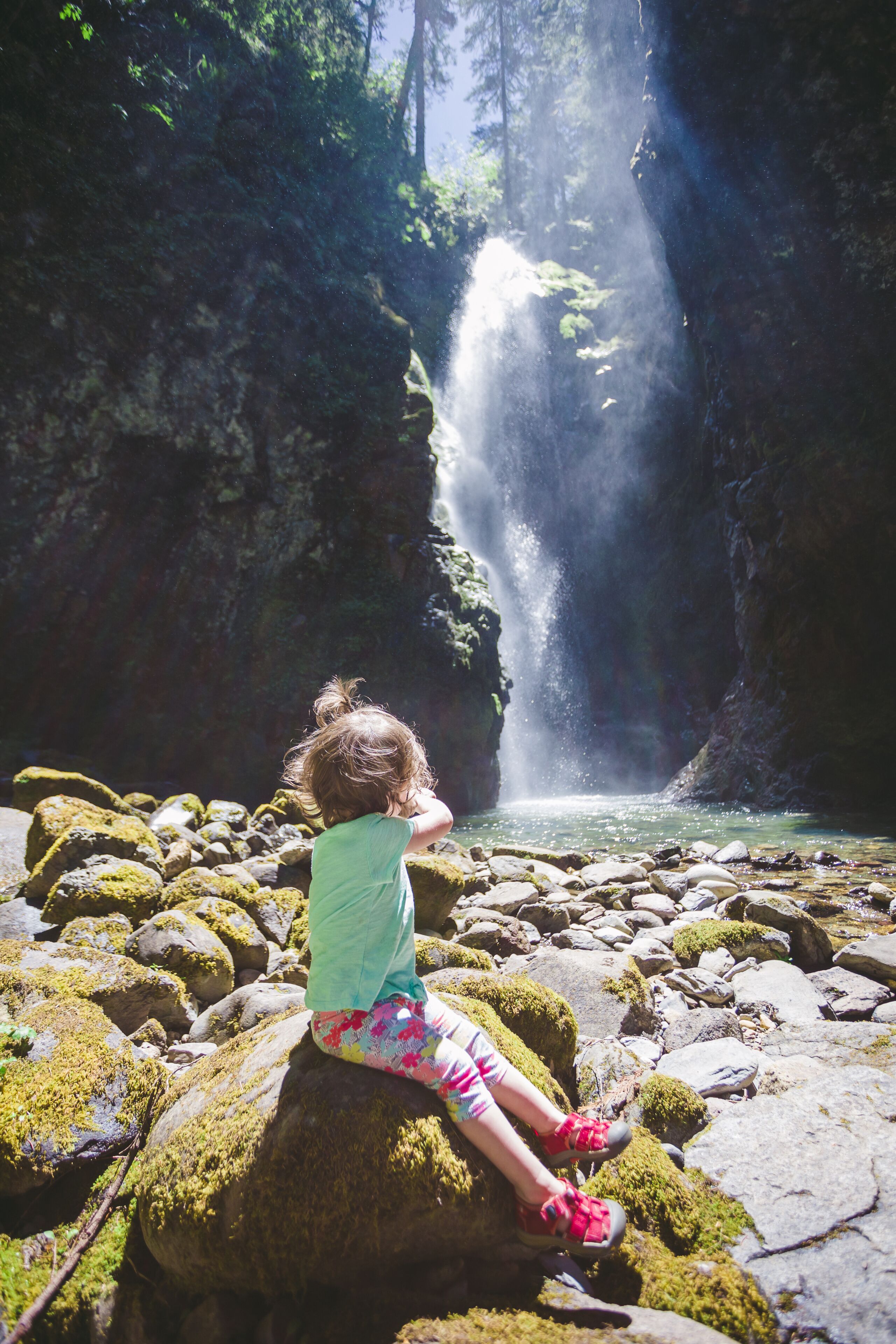 Criança sentada em uma rocha coberta de musgo perto de uma cascata.