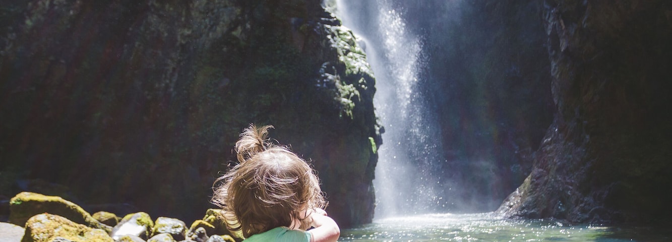 Criança sentada em uma rocha coberta de musgo perto de uma cascata.