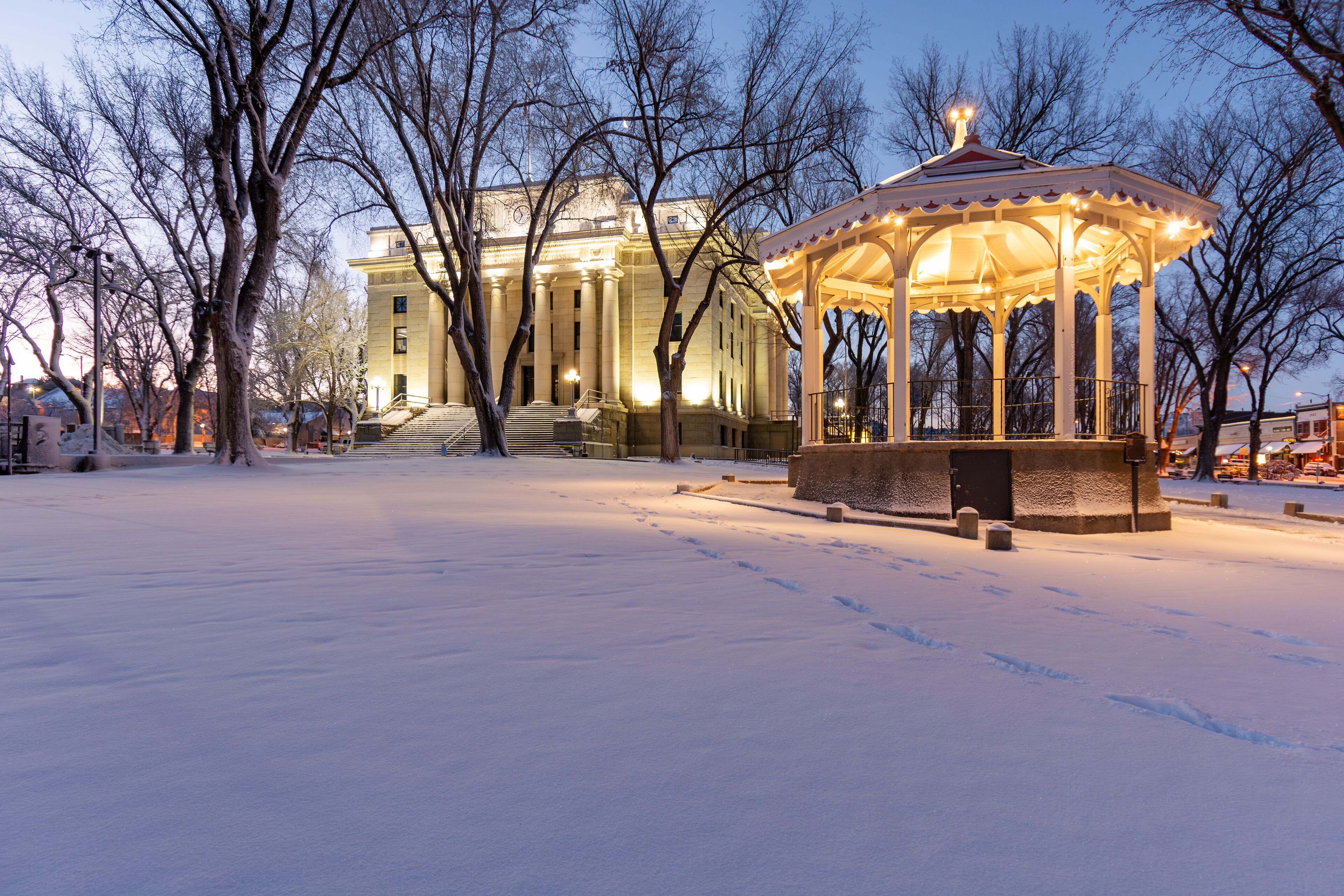 Um parque coberto de neve, com um coreto e um edifício clássico ao fundo.
