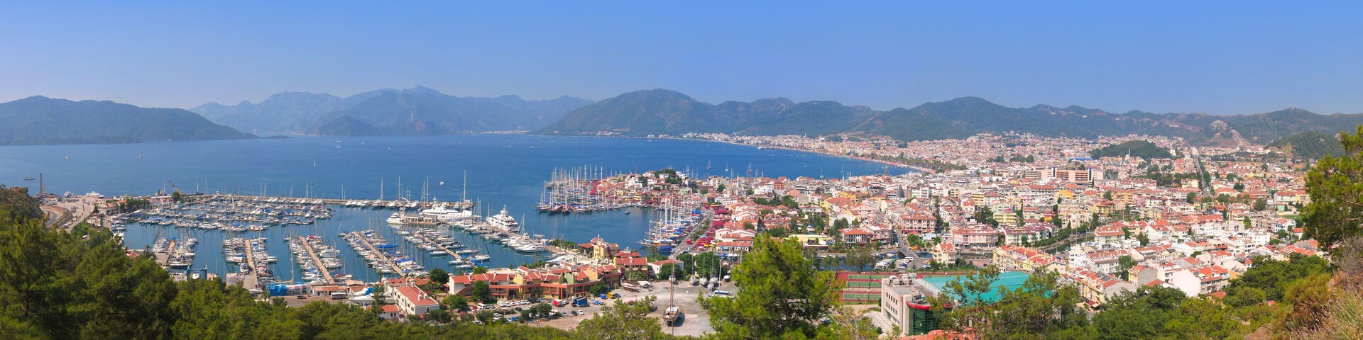 Uma cidade costeira com um porto repleto de barcos, rodeada por montanhas e uma área urbana densamente povoada.