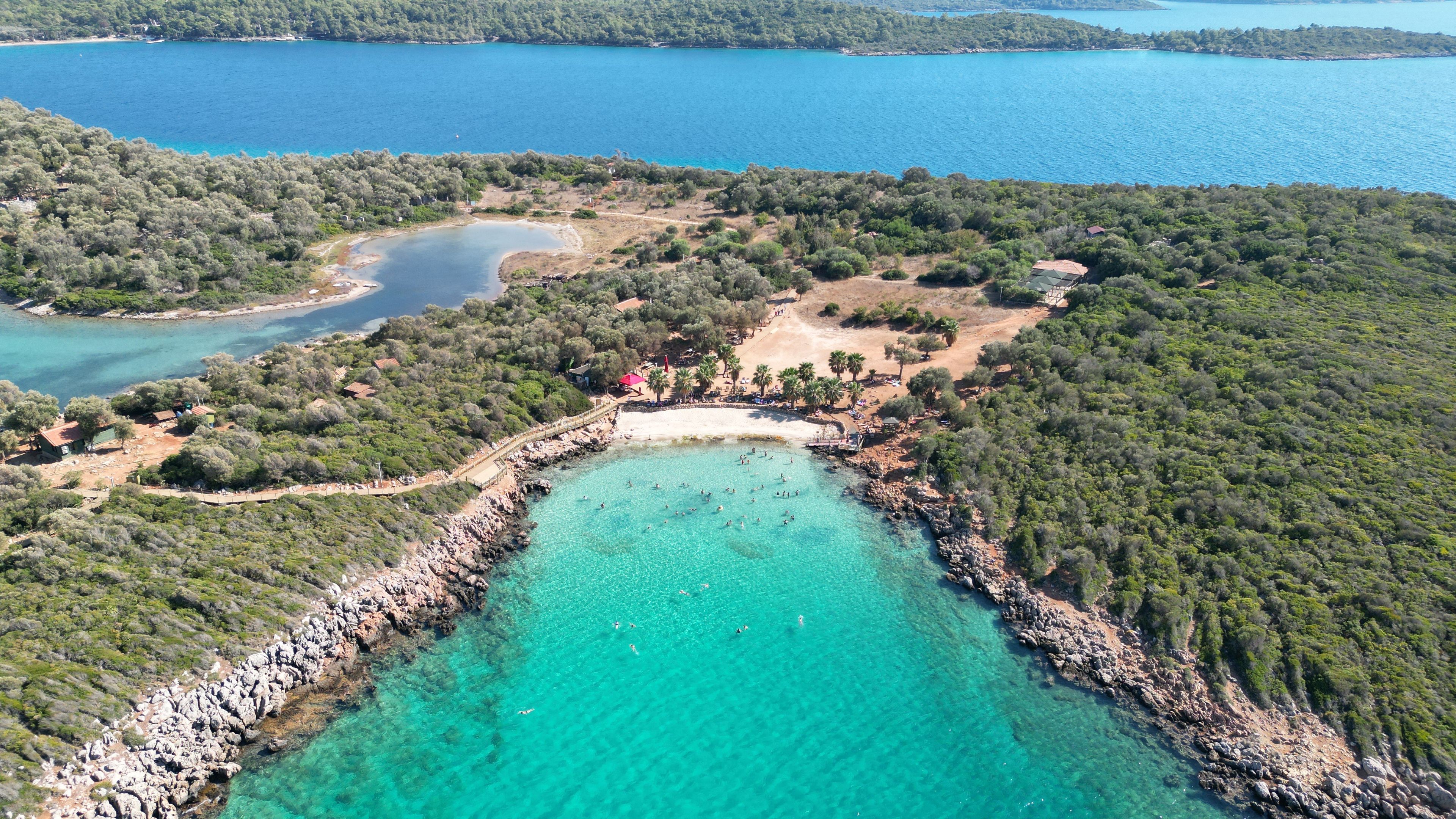 Vista aérea de uma área costeira com uma baía turquesa límpida, costa rochosa, floresta verde densa e uma praia de areia com pessoas.