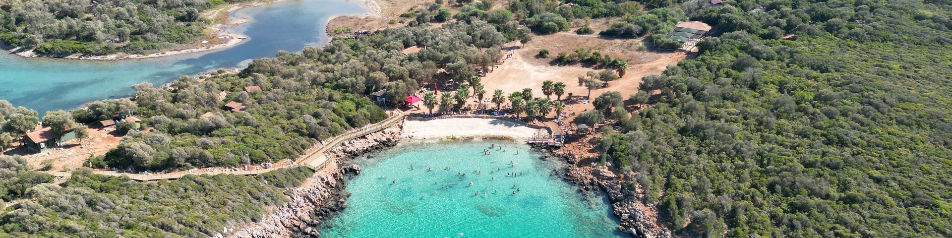 Vista aérea de uma área costeira com uma baía turquesa límpida, costa rochosa, floresta verde densa e uma praia de areia com pessoas.