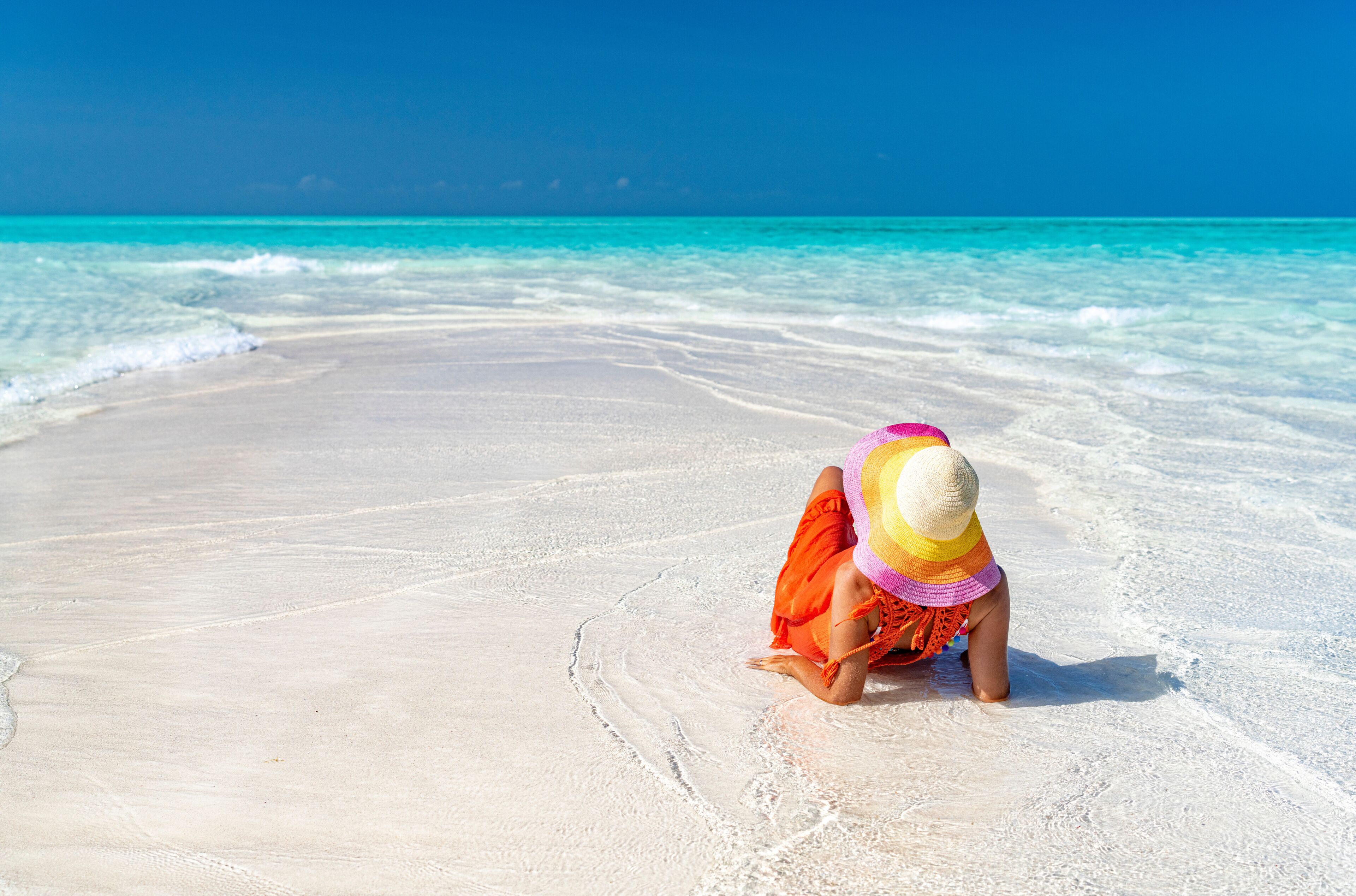 Uma praia de areia com água turquesa cristalina e uma pessoa usando um chapéu colorido e roupas laranja.