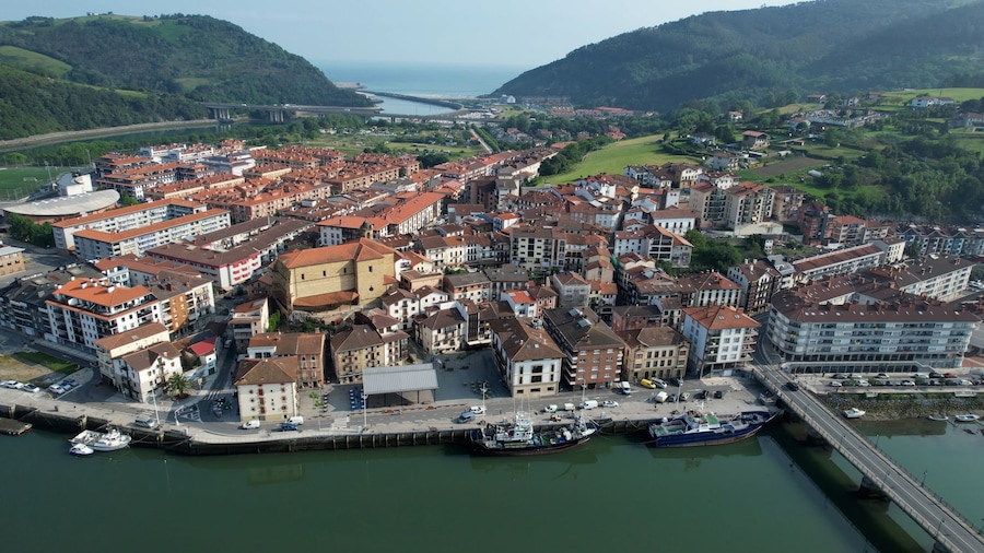 Vista aérea de uma cidade costeira com edifícios de telhados vermelhos, um porto de iates e uma ponte ao fundo.