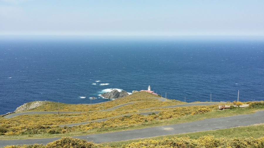 Paisagem costeira com um farol numa colina, uma estrada sinuosa e o oceano.