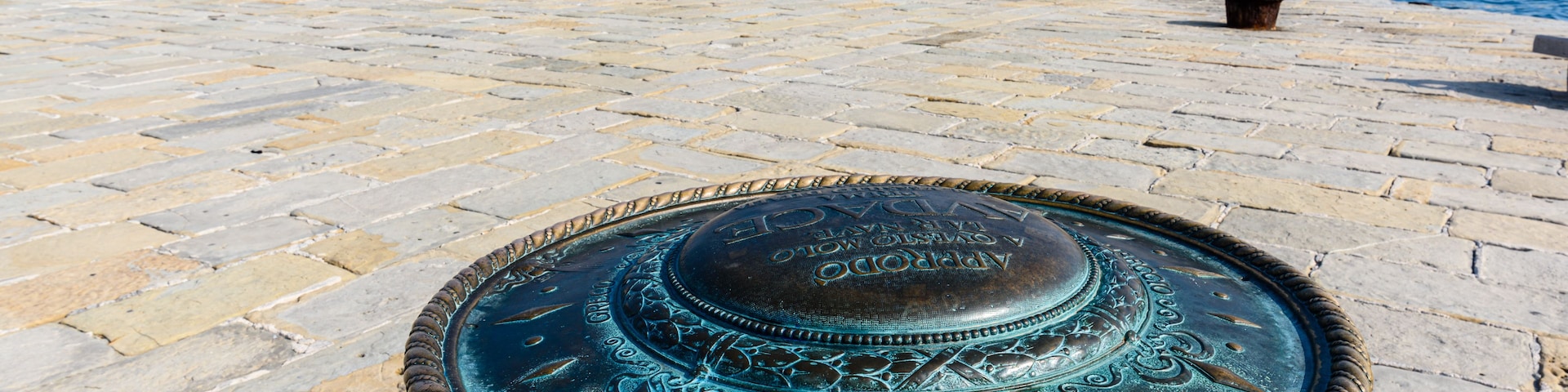 Um calçadão à beira-mar, com uma bacia de metal decorativa em primeiro plano e edifícios históricos ao fundo.