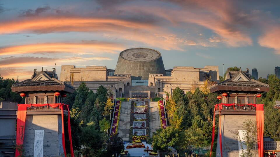 Um edifício tradicional chinês com uma estrutura circular central, rodeado por árvores e decorações vermelhas.