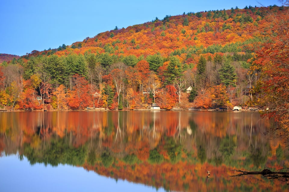 Um lago refletindo a folhagem outonal, com uma colina ao fundo e uma casa pequena perto da margem.